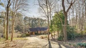 Uma casa com telhado inclinado, cercada por árvores e uma entrada com brita.