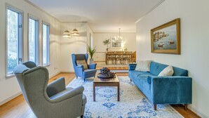 Uma sala de estar ampla com um sofá azul, poltronas cinzas e uma área de jantar ao fundo.