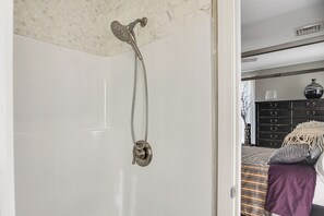 Casa de banho com chuveiro, parede branca e cama com colcha e almofadas riscadas.