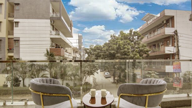 Varanda com duas cadeiras cinzas estofadas e uma pequena mesa, com vista para a rua com edifícios e árvores.