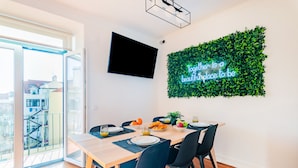 Uma área de refeições com mesa de madeira posta para quatro pessoas, cadeiras pretas, um letreiro de neon na parede e uma televisão montada acima da mesa.