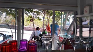 Uma área de bar com vista para a rua, uma mulher sentada à mesa e garrafas de vidro coloridas.