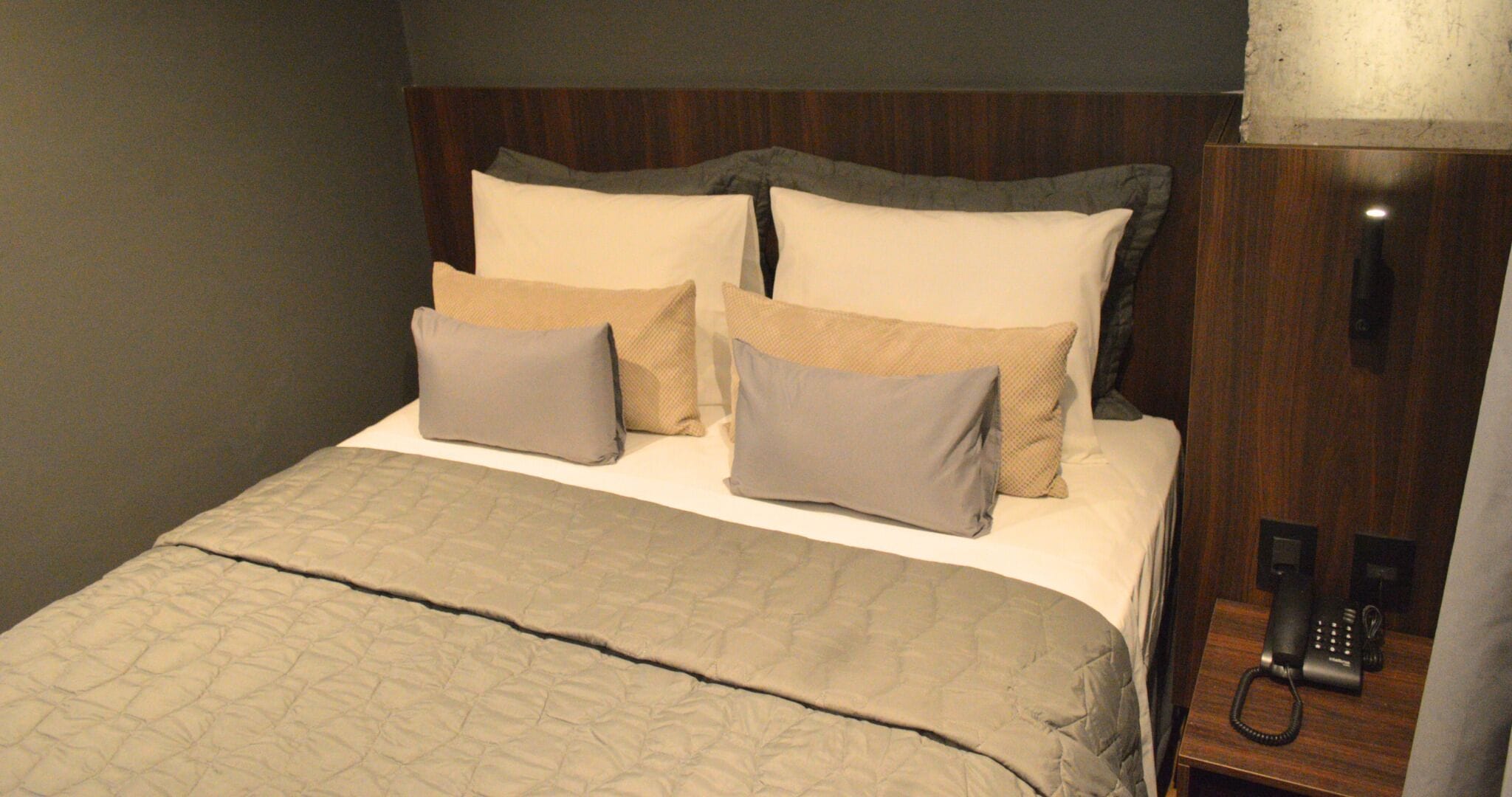 Quarto de hotel com cama, travesseiros, colcha acolchoada, cabeceira de madeira e telefone ao lado da cama.
