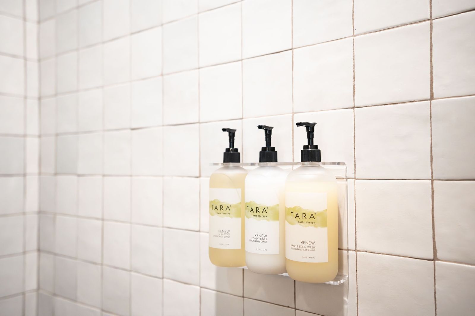 Três frascos de produtos TARA para banho sobre uma parede azulejada.