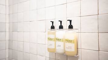 Três frascos de produtos TARA para banho sobre uma parede azulejada.