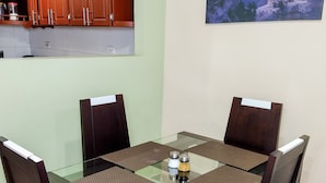Uma área de refeições com mesa de vidro, quatro cadeiras e um quadro de uma paisagem montanhosa na parede.
