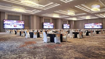 Uma sala de banquetes com fileiras de mesas e cadeiras, grandes telas exibindo uma programação e lustres elaborados.
