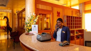 Balcão de recepção de hotel com recepcionista, placa de recepção, leitor de passaportes e arranjo de flores.