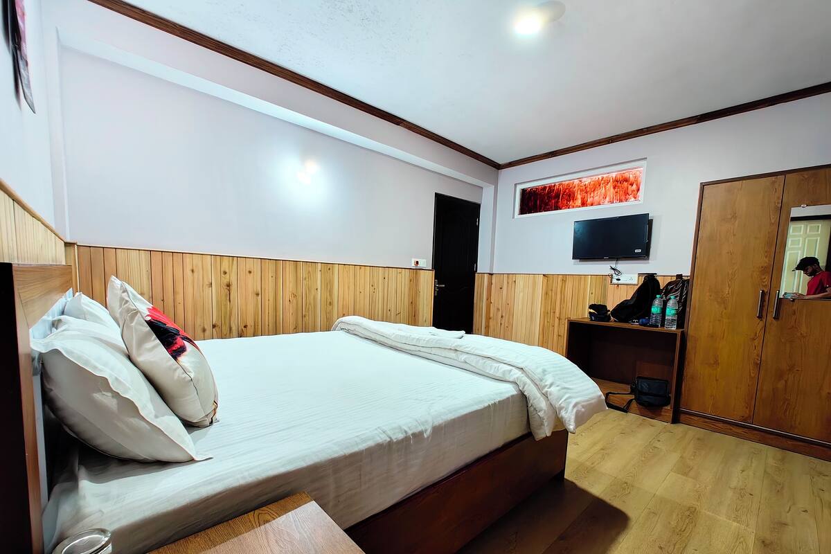 Quarto de hotel bem organizado com cabeceira de madeira, cama com roupa de cama branca, uma pequena mesa de madeira com um frasco de vidro, TV fixada na parede e guarda-roupa com espelho.