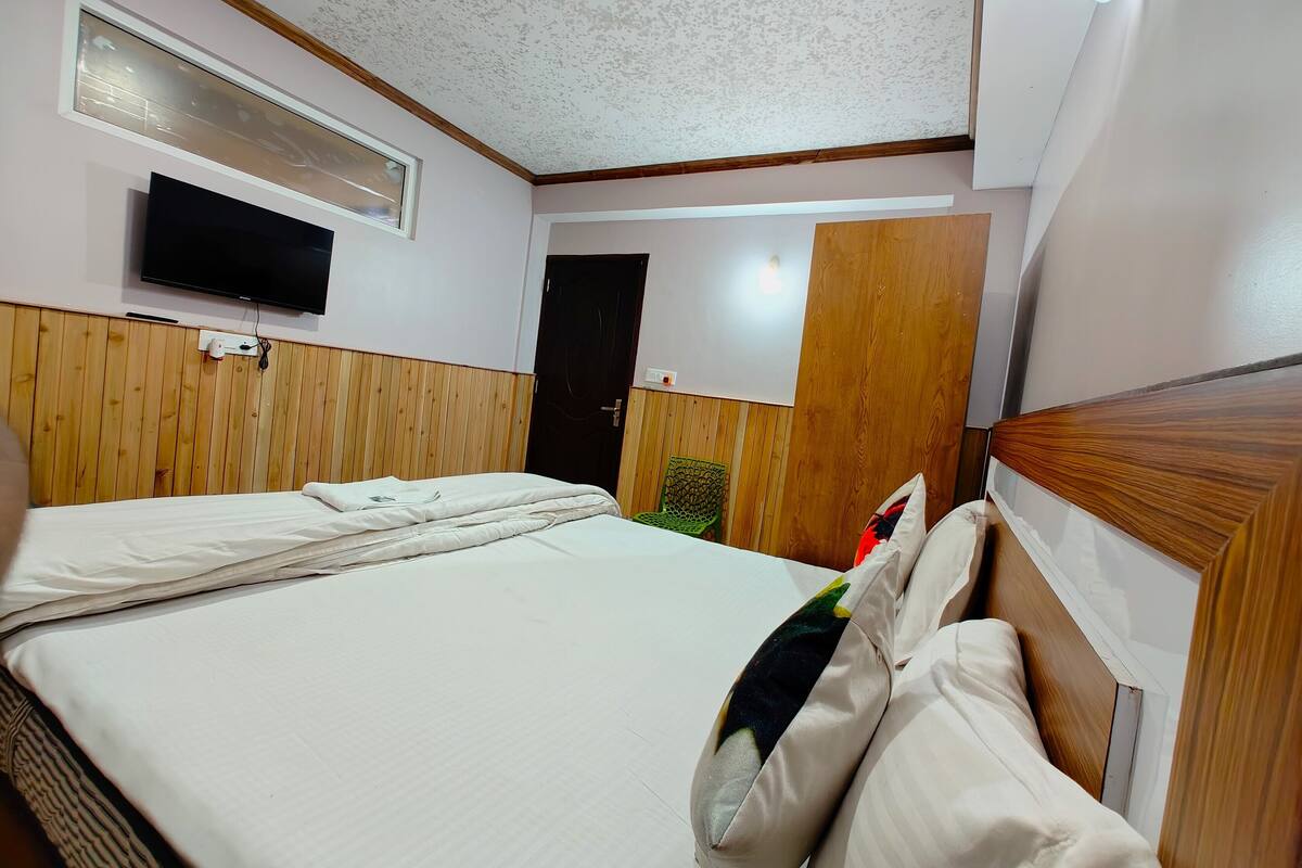 Quarto de hotel com cama, televisão fixada na parede e cabeceira de madeira.