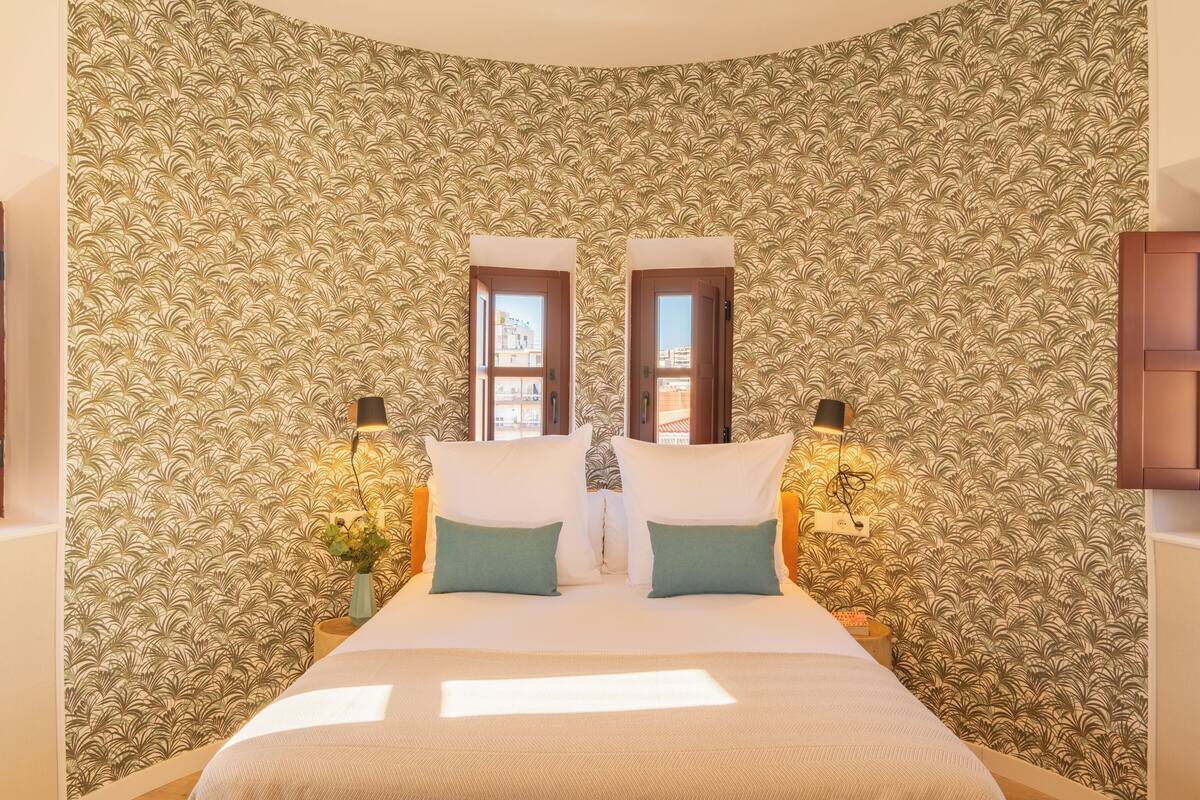 Quarto com papel de parede estampado, duas janelas, cama com almofadas, abajures e mesa de cabeceira com um vaso.