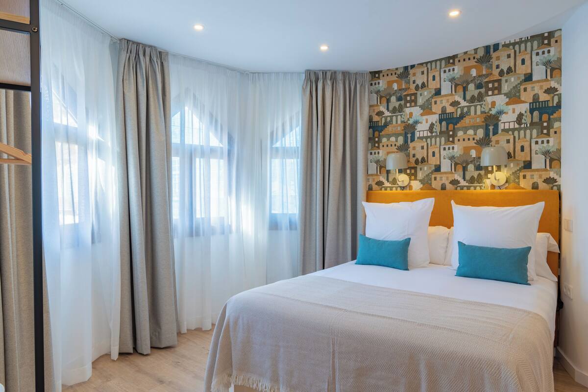 Quarto de hotel com uma cama grande, parede estampada e cortinas leves.