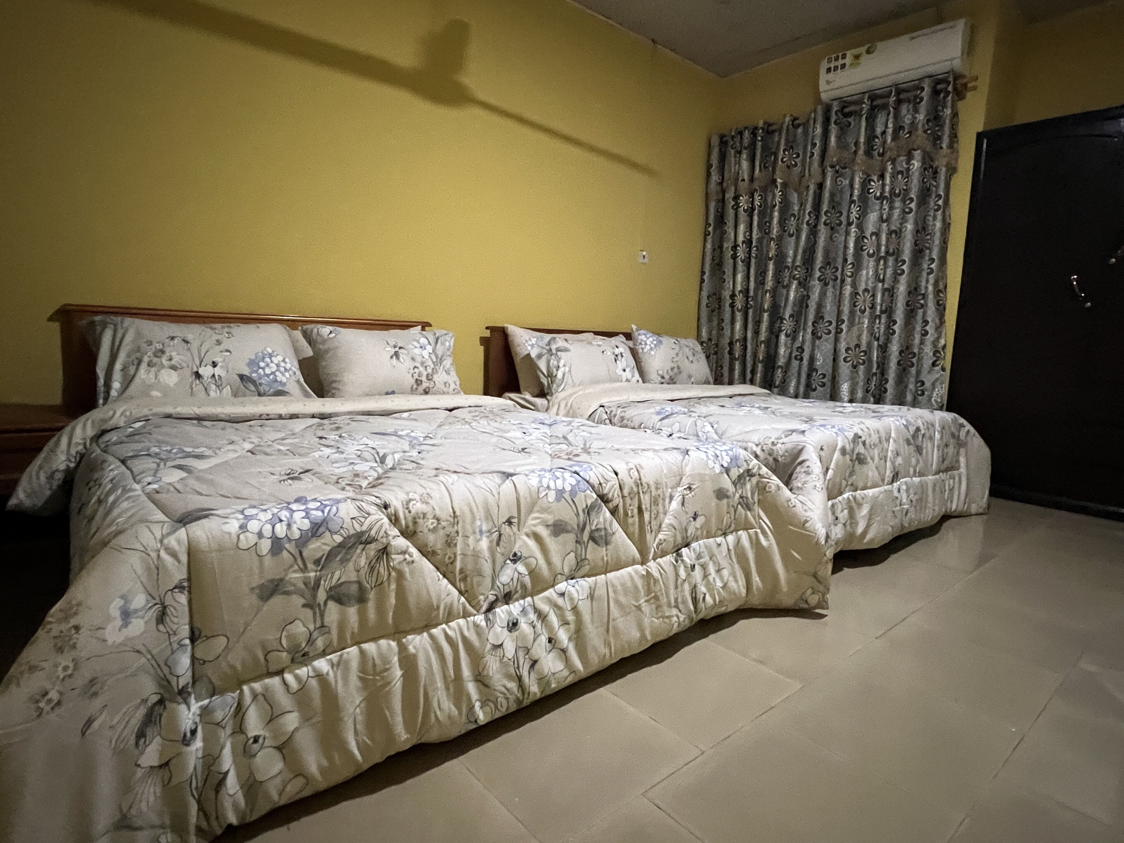 Quarto com duas camas, roupa de cama florida, janela com cortinas estampadas e ventilador de teto.
