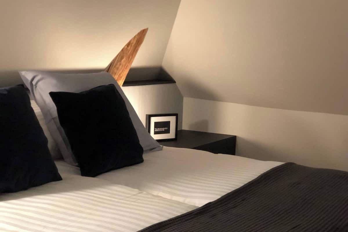 Uma cama bem arrumada com roupa de cama branca e duas almofadas escuras, uma pequena mesa de cabeceira com um quadro emoldurado e um objeto decorativo de madeira na parede.