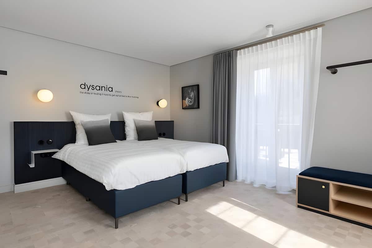 Quarto moderno com cama, mesas de cabeceira, um banco e luminária fixada na parede.