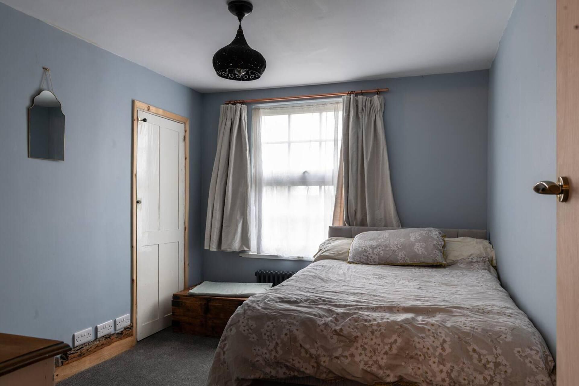 Um quarto com cama, janela com cortinas, porta, radiador e luminária pendente.
