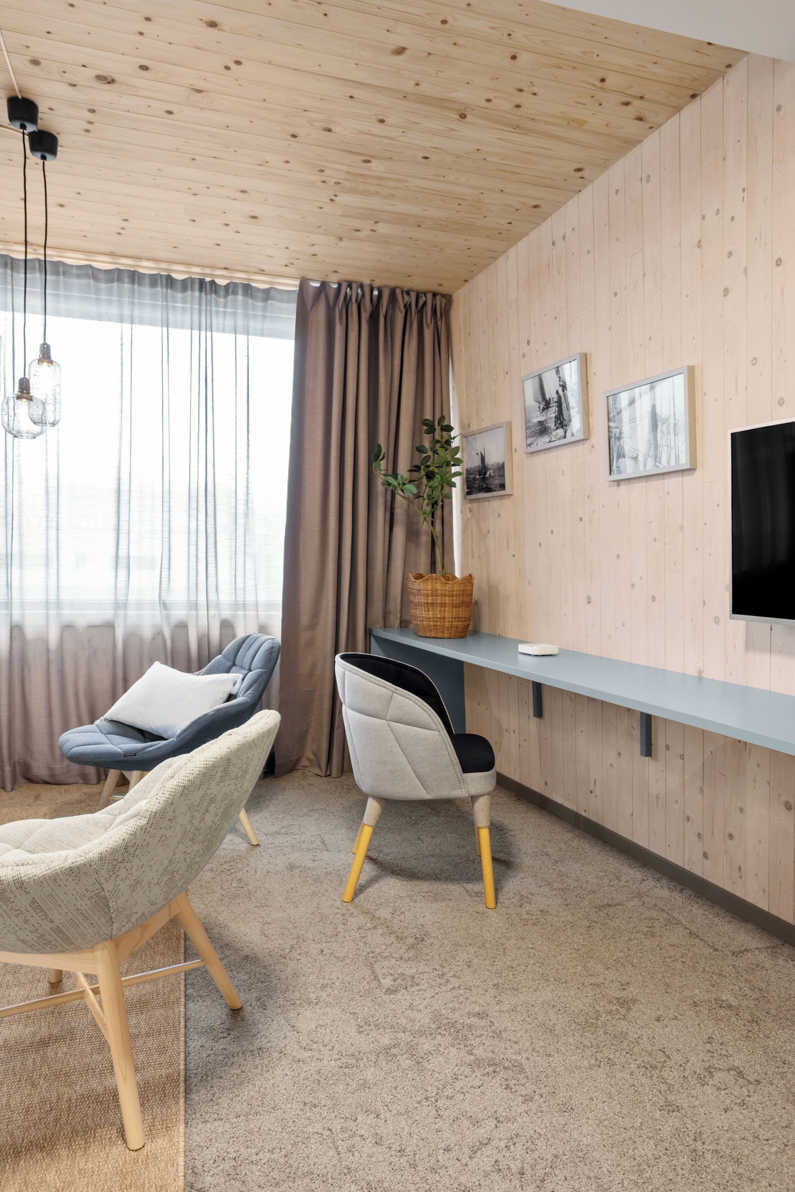 Quarto com teto de madeira, uma janela grande com cortinas, televisão fixada na parede e duas cadeiras.