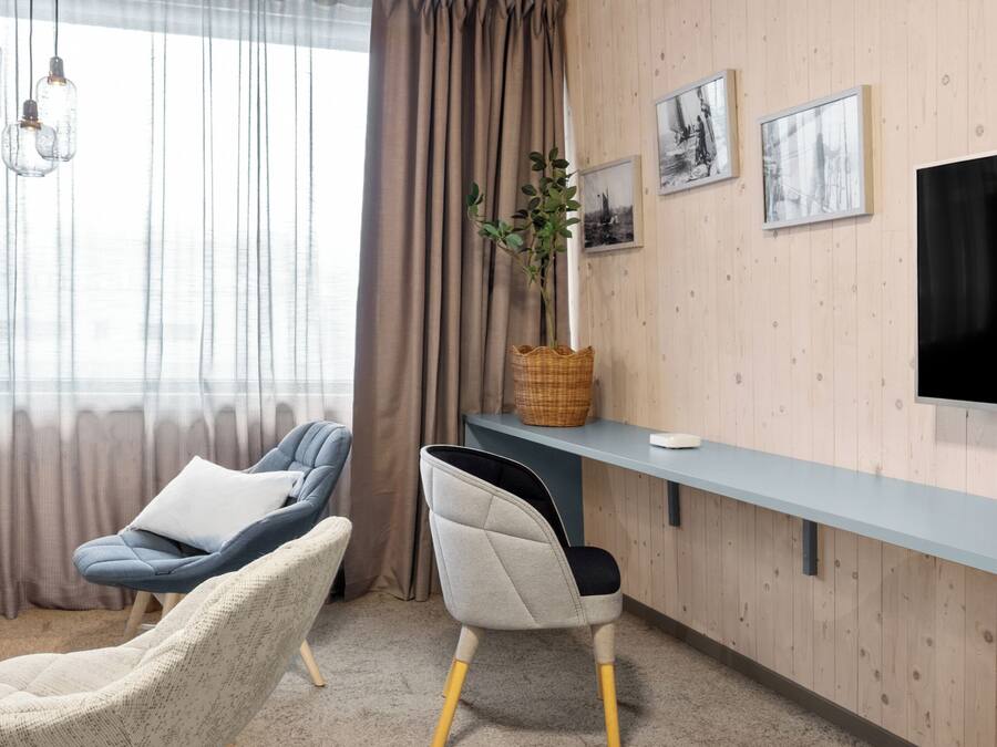Quarto com teto de madeira, uma janela grande com cortinas, televisão fixada na parede e duas cadeiras.
