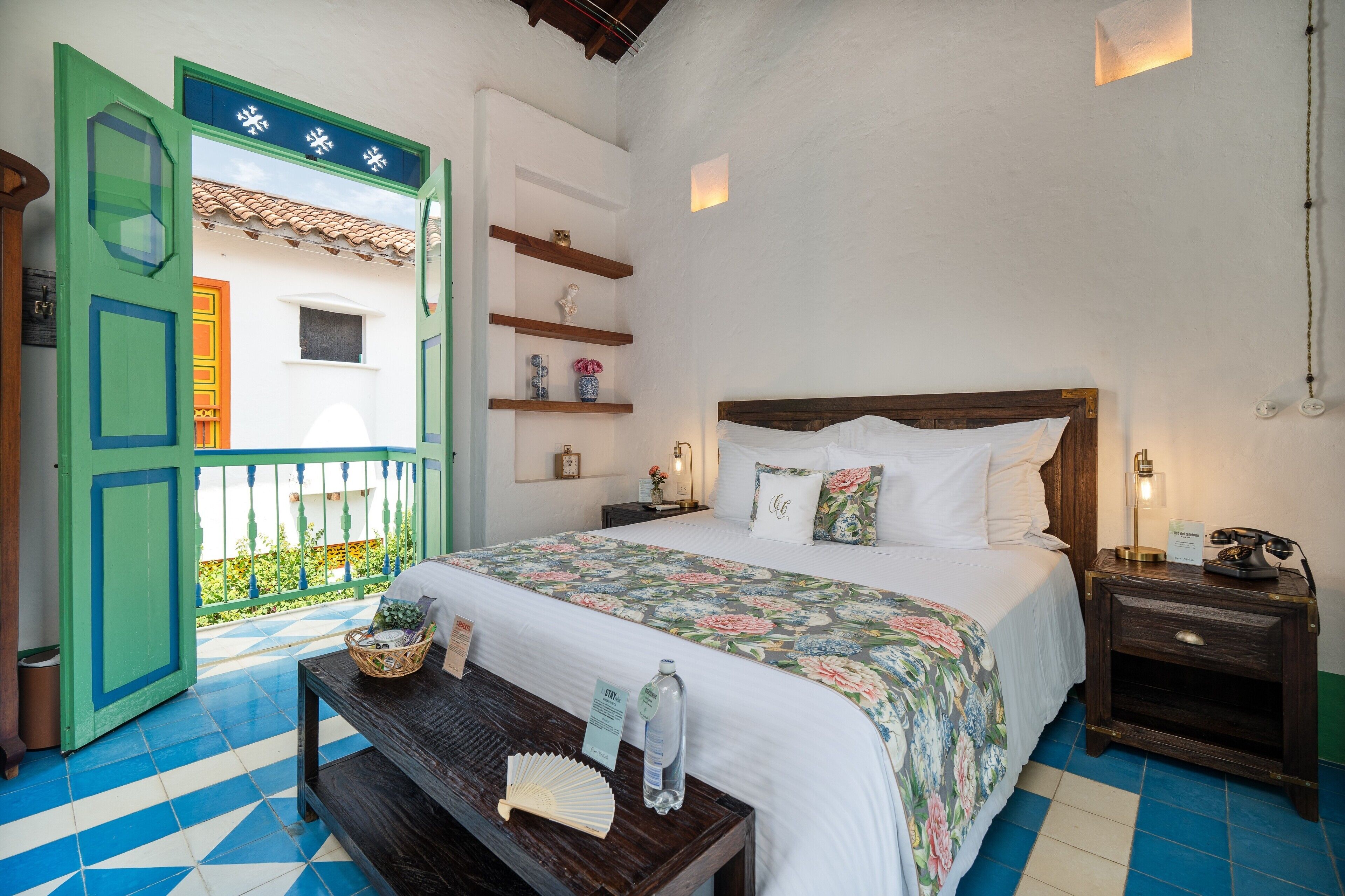 Um quarto com uma cama, mesas de cabeceira, um criado-mudo, uma janela e uma porta aberta para uma varanda.