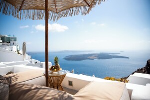 Um terraço à beira-mar com arquitetura branca, uma mesa de vime com frutas e vista desimpedida para o mar e ilhas.