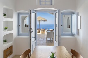 Uma porta aberta dá para um terraço com vista para o mar, uma mesa de madeira e cadeiras dentro de um quarto com paredes brancas e prateleiras com plantas.