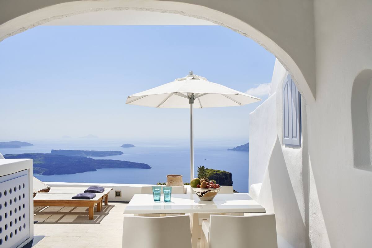 Uma varanda com guarda-sol branco, uma mesa com uma cesta de frutas e vista para o mar.