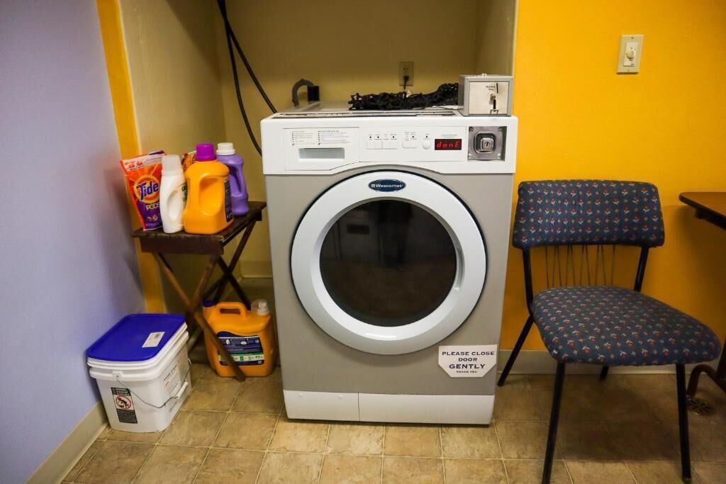 Uma lavanderia compacta com máquina de lavar e secar frontal, uma cadeira e diversos produtos de limpeza.