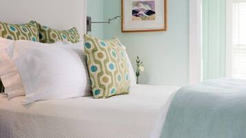 Cama bem arrumada com almofadas brancas e estampadas, um cobertor de linho azul claro e uma obra de arte emoldurada na parede.