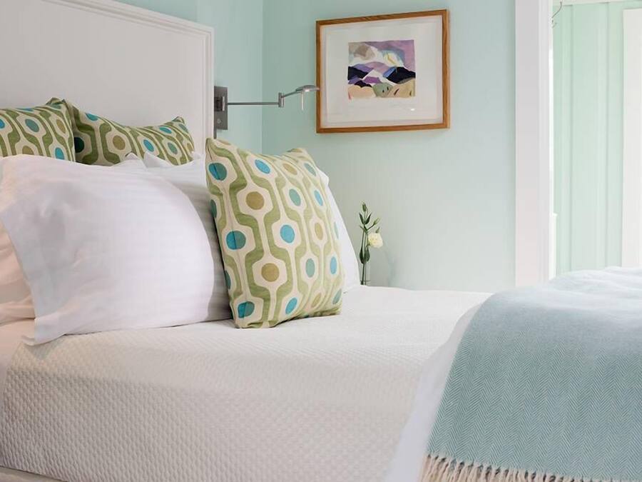 Cama bem arrumada com almofadas brancas e estampadas, um cobertor de linho azul claro e uma obra de arte emoldurada na parede.
