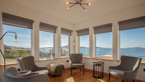 Quarto amplo com grandes janelas com vista para o mar, um lustre moderno e assentos confortáveis.