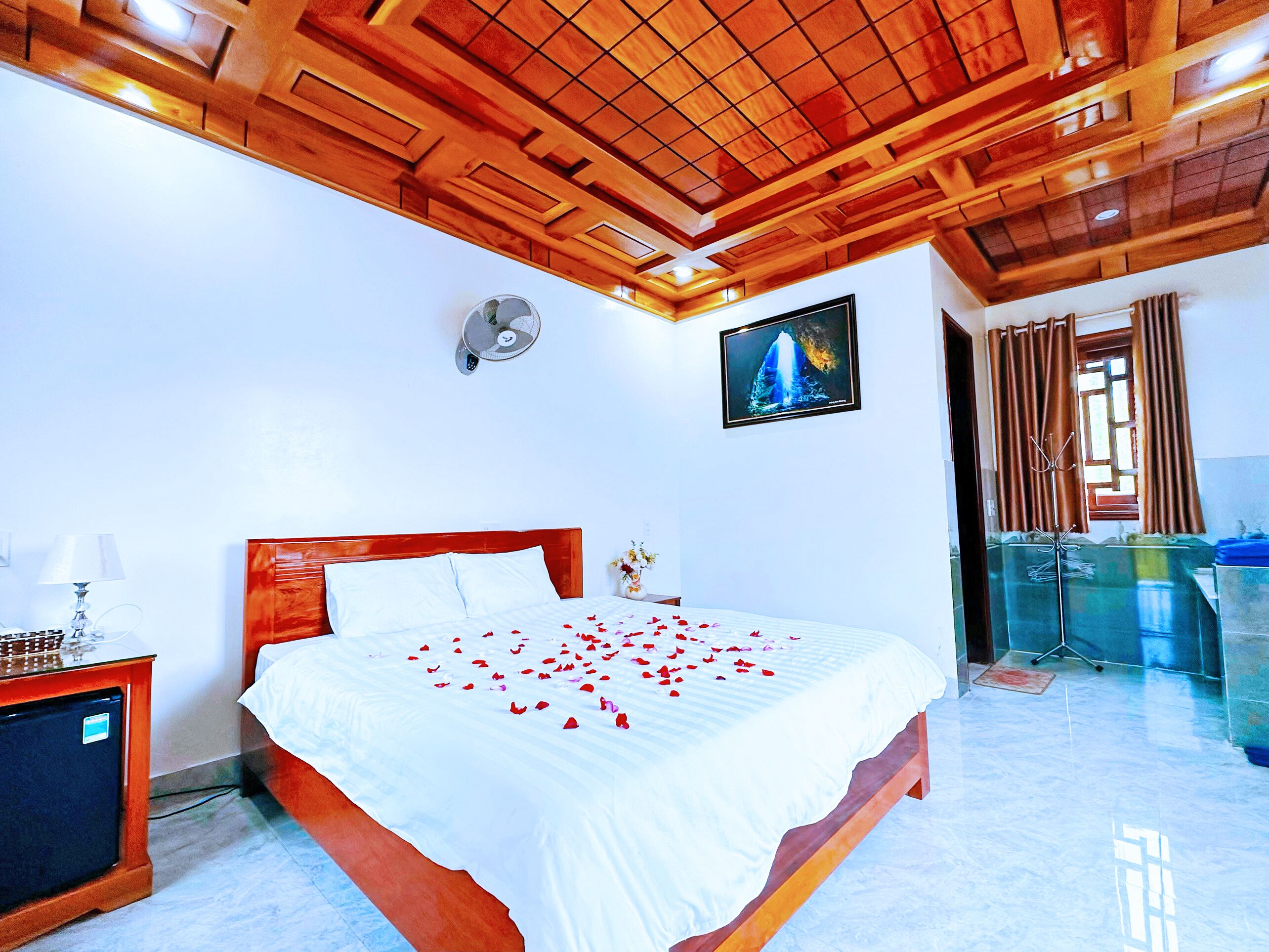 Quarto de hotel com teto de madeira, cama com pétalas de rosa, ventoinha na parede e uma moldura com imagem.