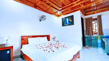 Quarto de hotel com teto de madeira, cama com pétalas de rosa, ventoinha na parede e uma moldura com imagem.