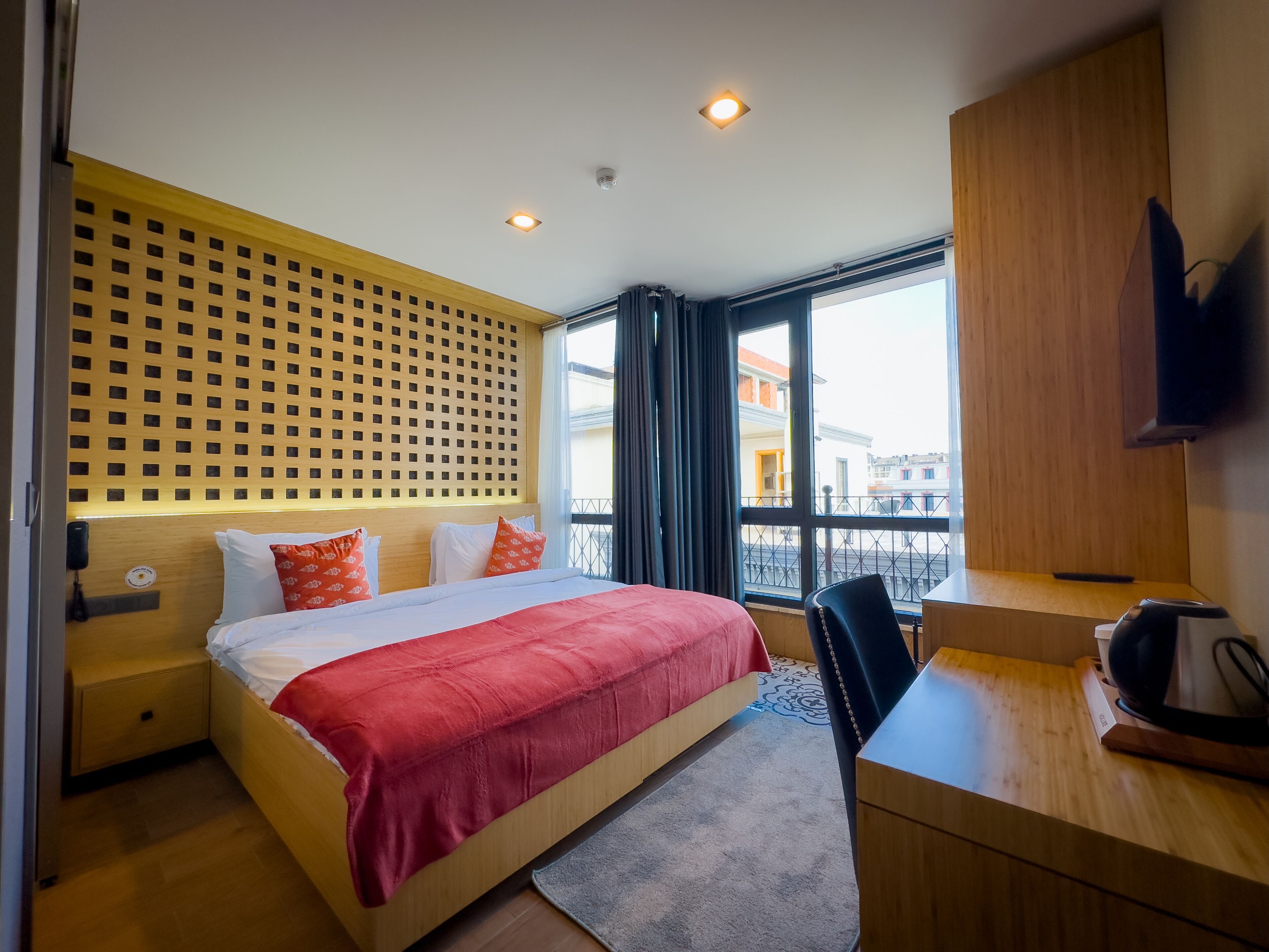 Quarto de hotel com cama, escrivaninha, cadeira, televisão e janela com vista para edifícios.
