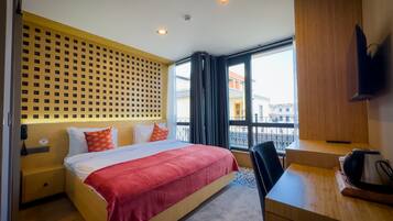 Quarto de hotel com cama, escrivaninha, cadeira, televisão e janela com vista para edifícios.