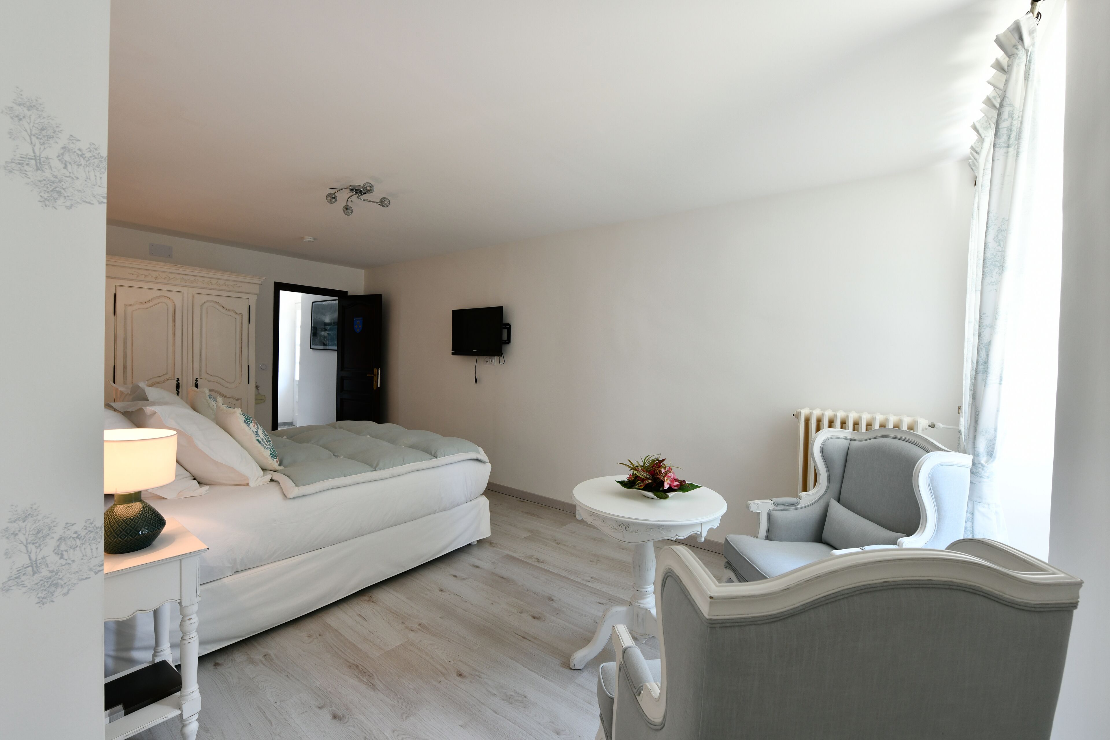 Quarto com cama, mesa de cabeceira, luminária, cadeira e uma pequena mesa com flores.