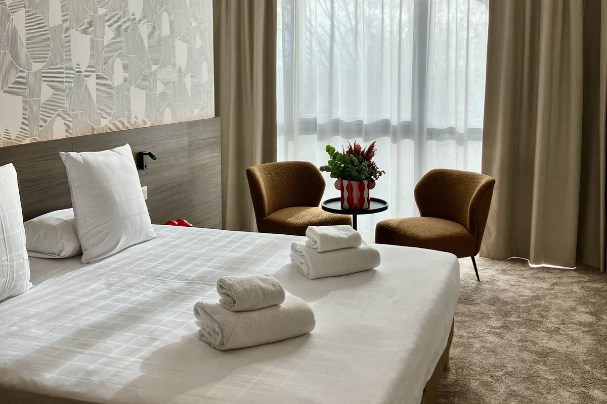 Quarto de hotel com uma cama grande, duas poltronas, uma mesinha com um vaso de flores e uma janela com cortinas.