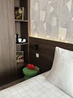Um quarto com cama, mesa de cabeceira, prateleira com livros, parede decorativa e papel de parede estampado.