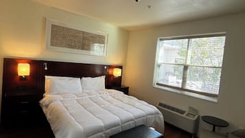Quarto de hotel com cama grande, abajures, um banco, uma mesinha, uma janela e uma câmera de segurança instalada no teto.