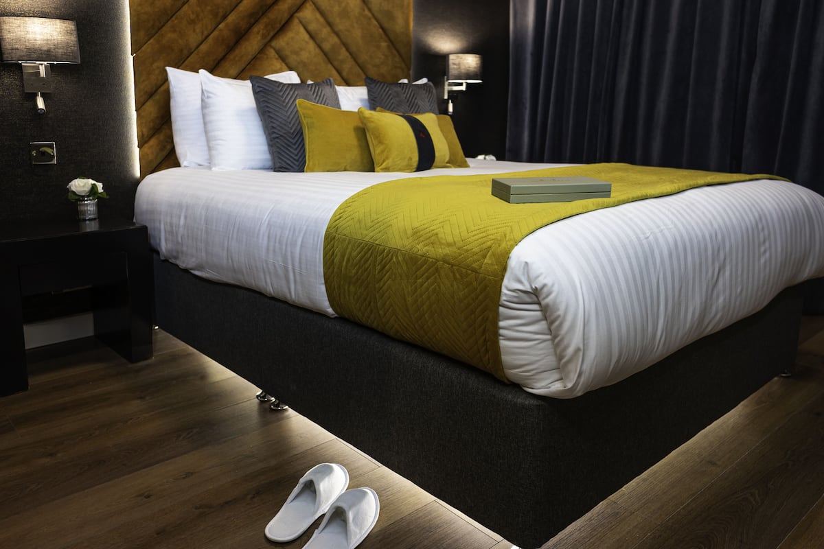 Quarto de hotel moderno com uma cama grande, uma mesa de cabeceira com abajur e um par de chinelos no chão.