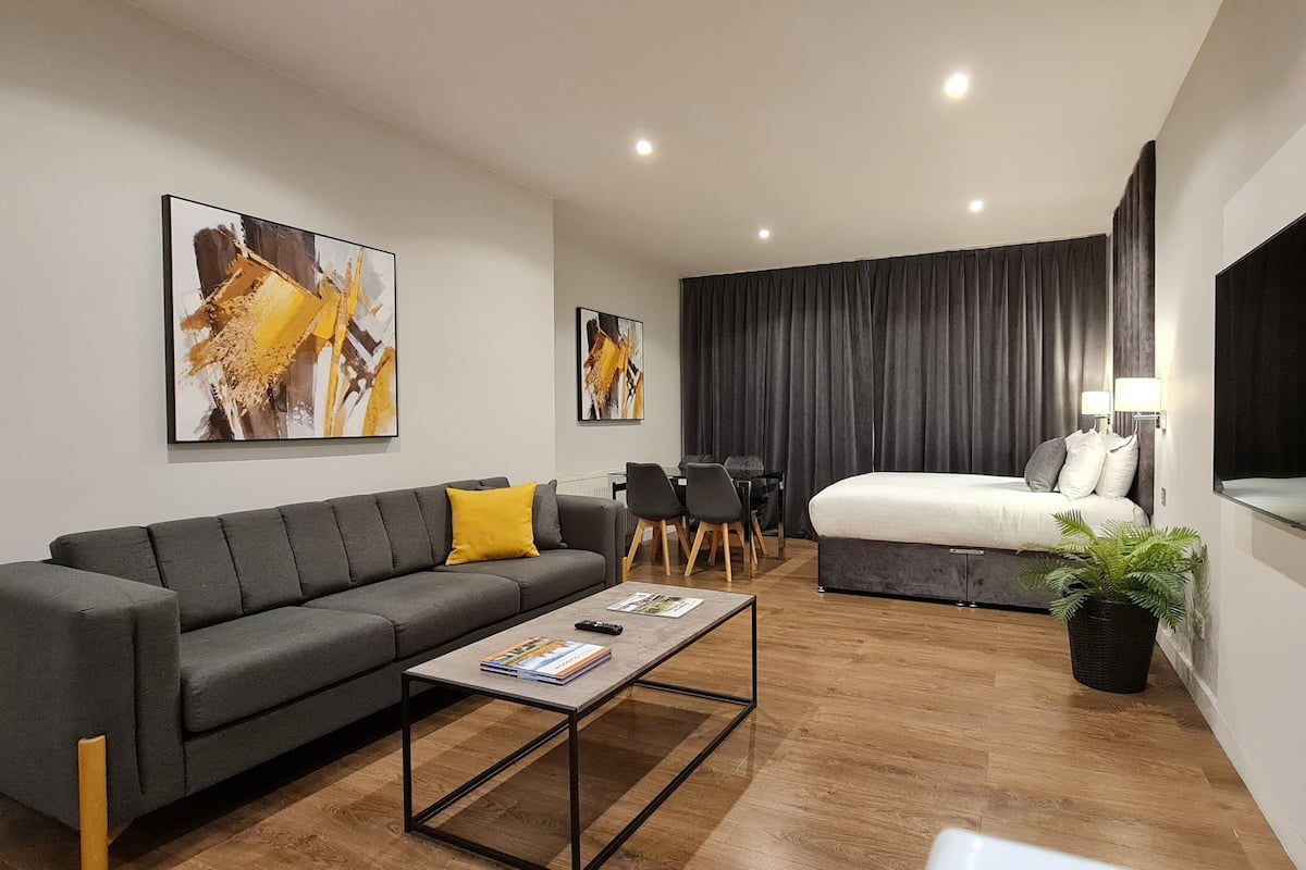 Quarto de hotel moderno com sofá cinza, mesa de centro pequena, área de refeições com cadeiras, cama com cabeceira preta e um espelho grande.