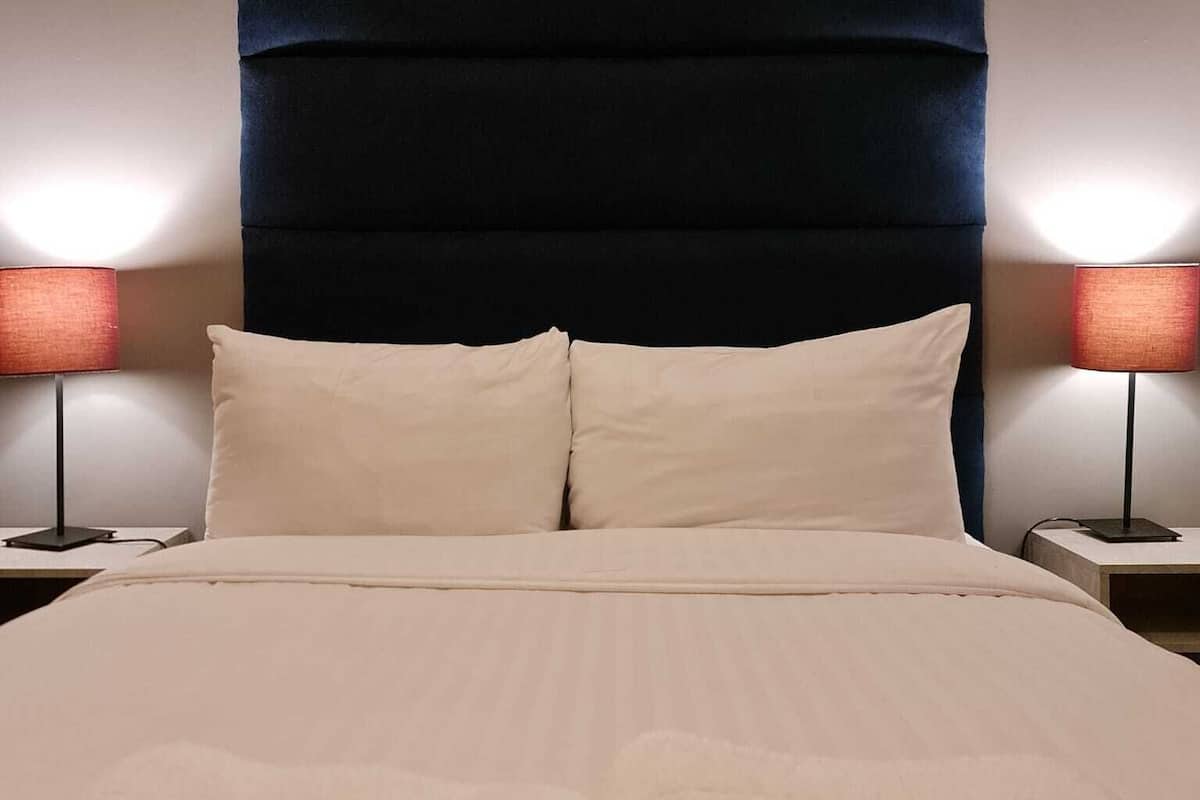 Quarto de hotel com uma cama grande, duas luminárias de cabeceira e um painel de cabeceira escuro.