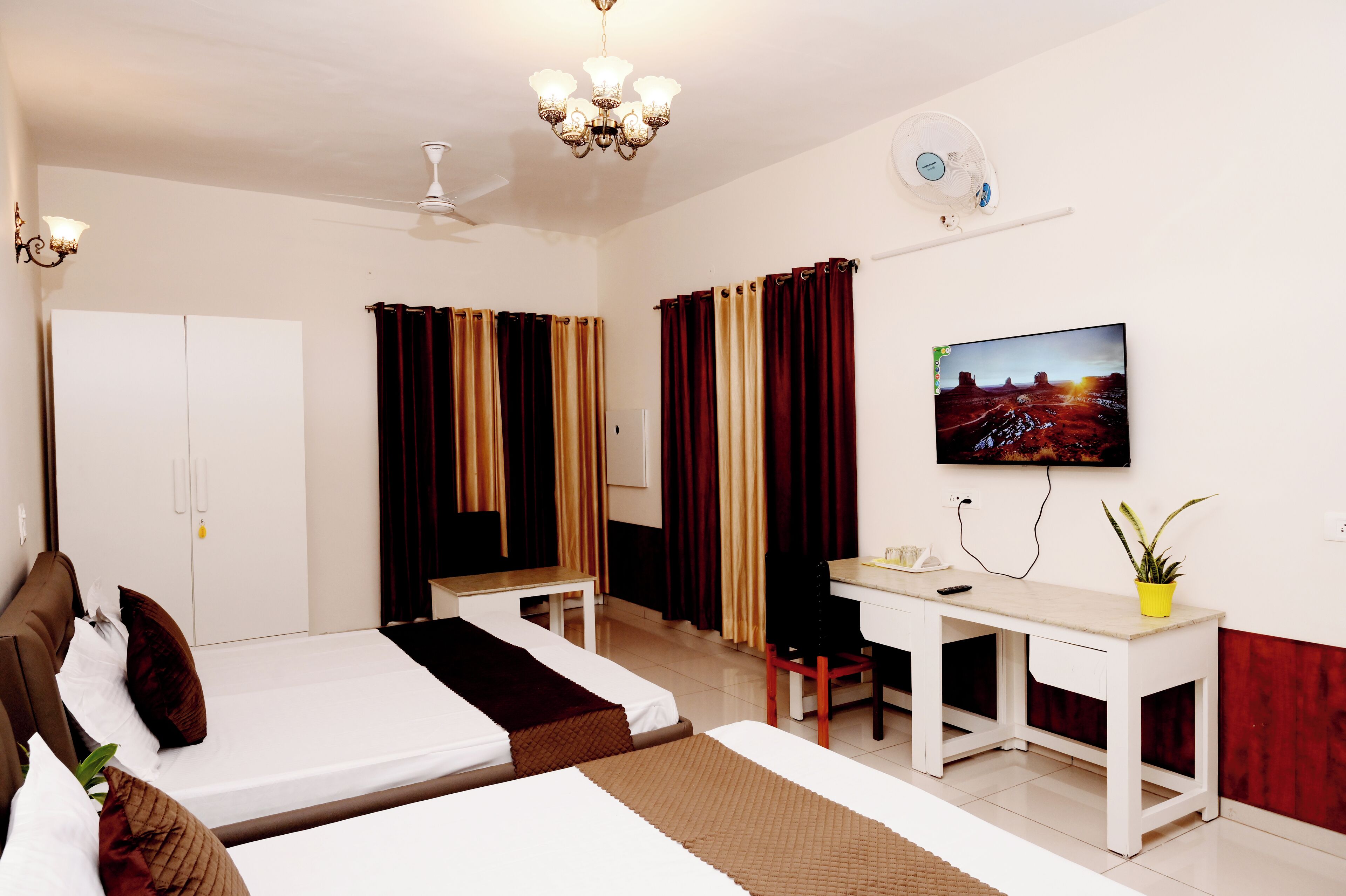Quarto de hotel com duas camas, uma escrivaninha, uma cadeira, uma televisão e um ventilador de teto.