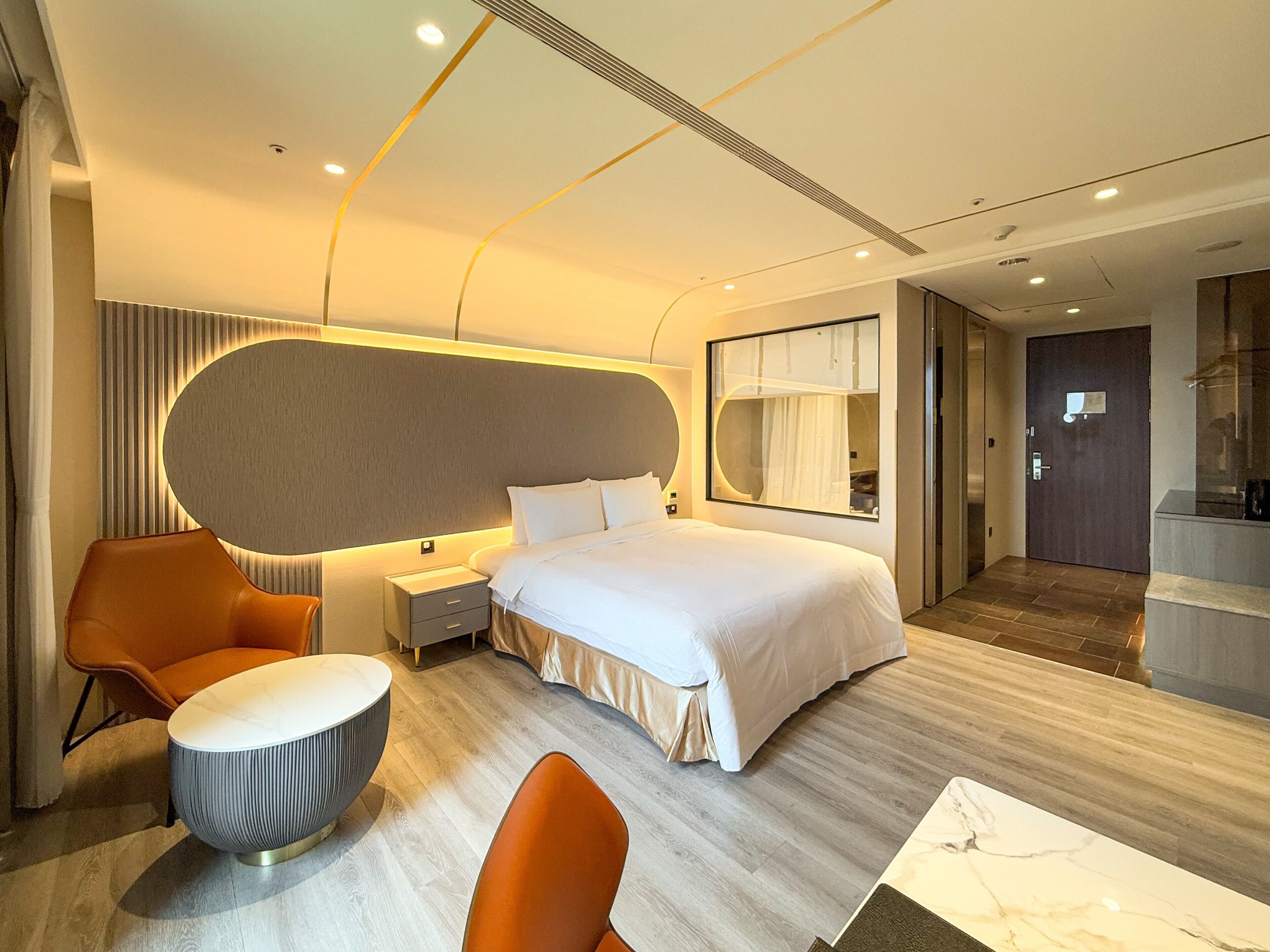 Quarto de hotel moderno com cama grande, mesa lateral redonda, poltrona laranja, espelho e piso de madeira.