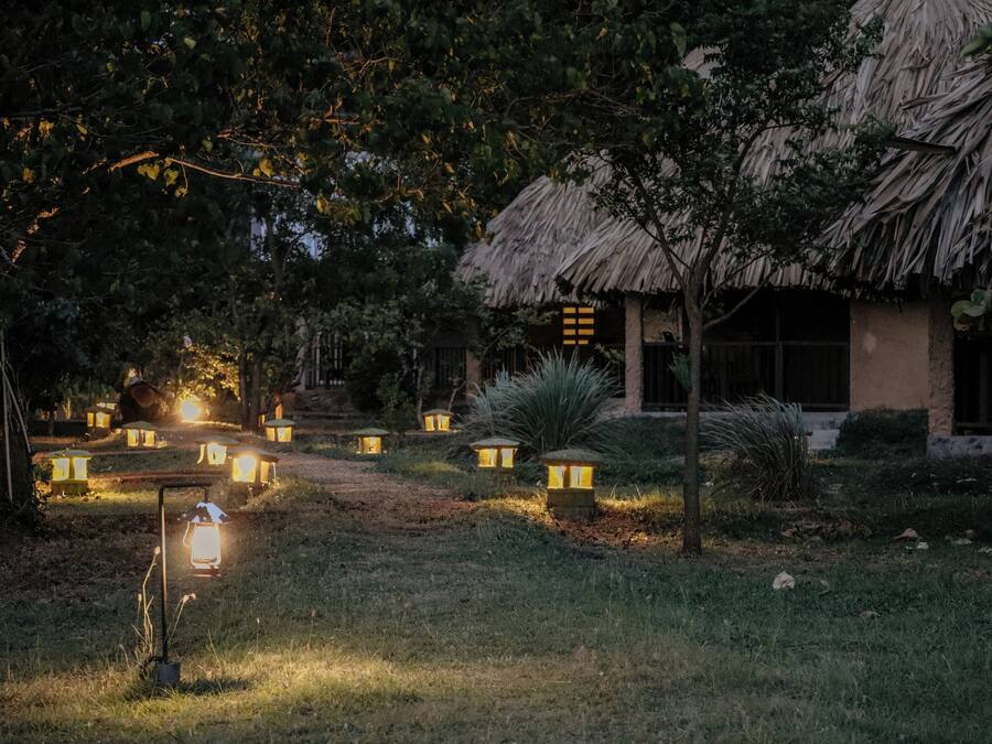 Uma área externa rústica com cabanas de telhado de palha, lanternas e um caminho.