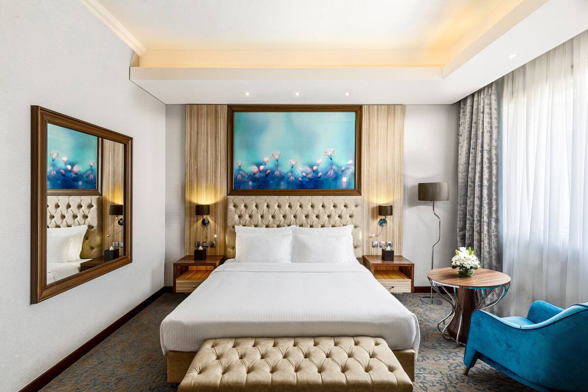 Quarto de hotel com uma cama grande, cabeceira estofada, uma mesa redonda com um vaso de flores, uma poltrona azul e duas luminárias fixadas na parede.