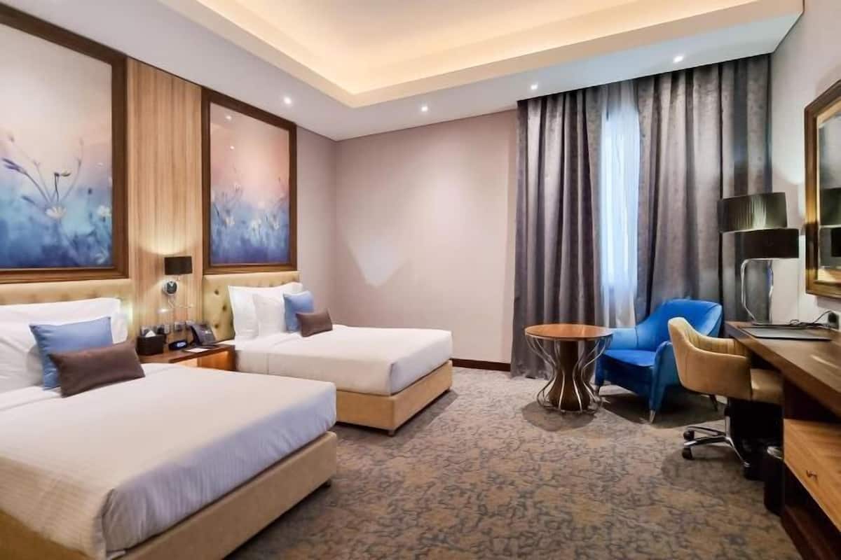 Quarto de hotel com duas camas, uma escrivaninha, uma cadeira, uma mesinha e uma janela grande com cortinas.