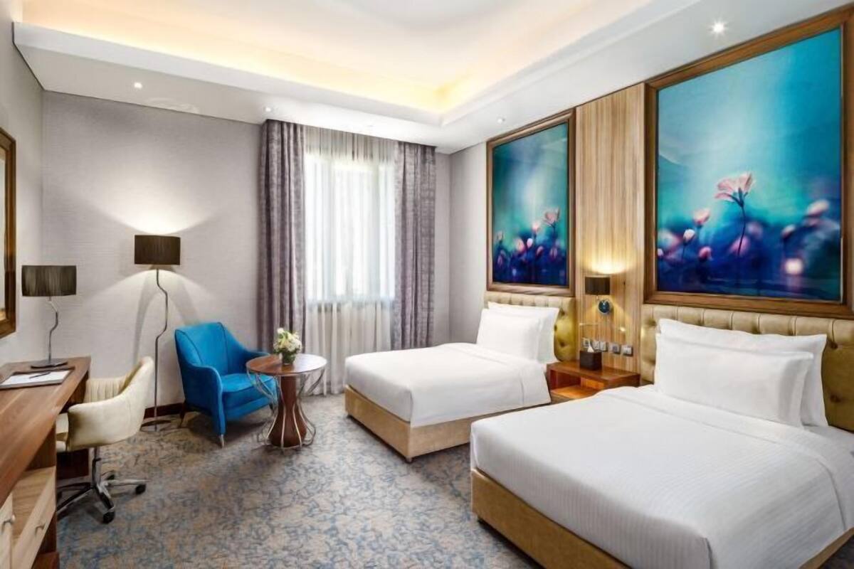 Quarto de hotel com duas camas, uma escrivaninha, uma cadeira, uma luminária e grandes pinturas florais nas paredes.