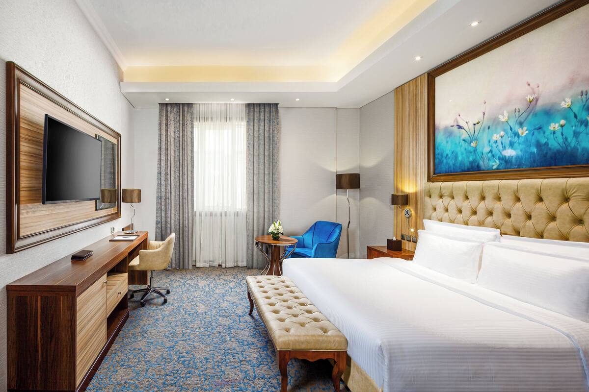 Quarto de hotel com uma cama grande, uma escrivaninha com televisão de tela plana, uma cadeira, um banco e um quadro na parede.