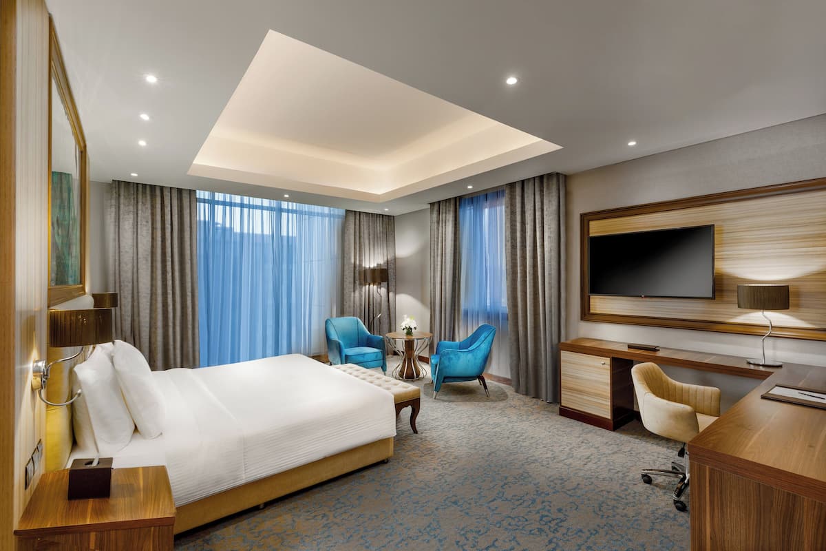 Quarto de hotel com uma cama grande, duas poltronas azuis, uma escrivaninha, uma TV e uma luminária no teto.