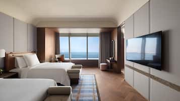 Quarto de hotel moderno com duas camas, uma escrivaninha, televisão e uma grande janela com vista para o mar.